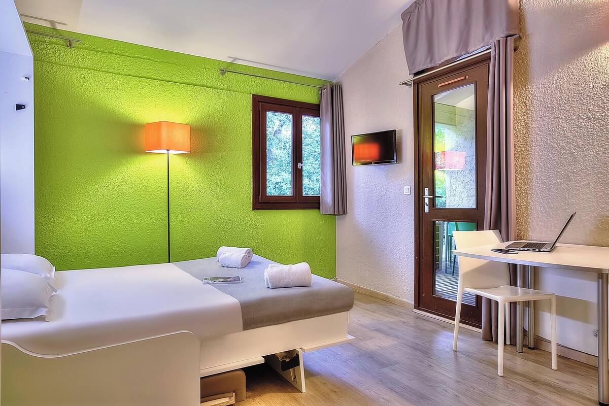 Quarto com cama, escrivaninha, cadeira, televisão e janela com cortinas.