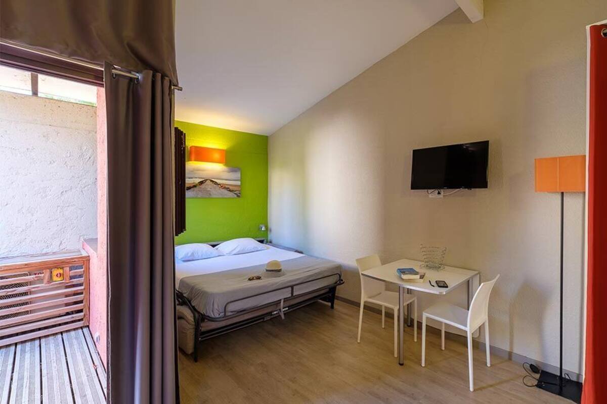 Um quarto pequeno e bem iluminado, com uma cama, uma mesa com cadeiras, uma TV fixada na parede e uma janela com cortinas.