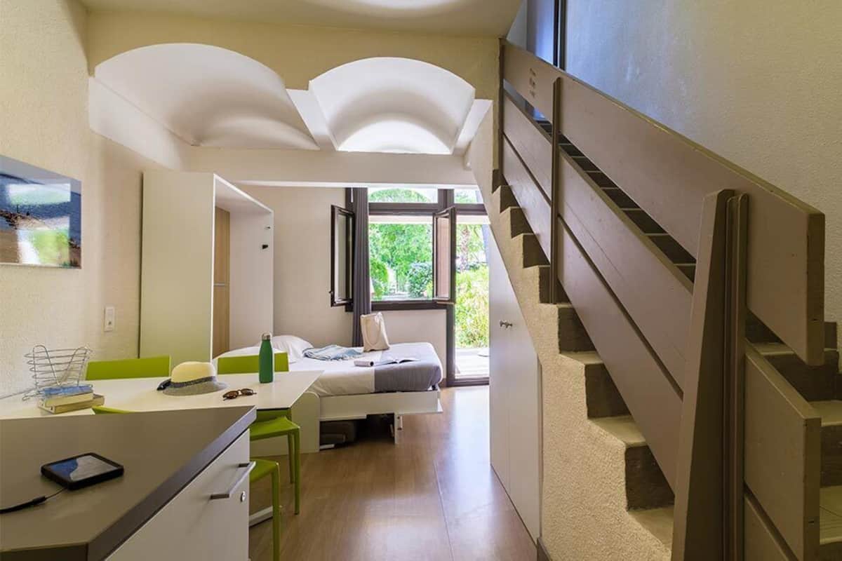 Quarto de hotel moderno com escada, cama, escrivaninha e vista para o verde lá fora.
