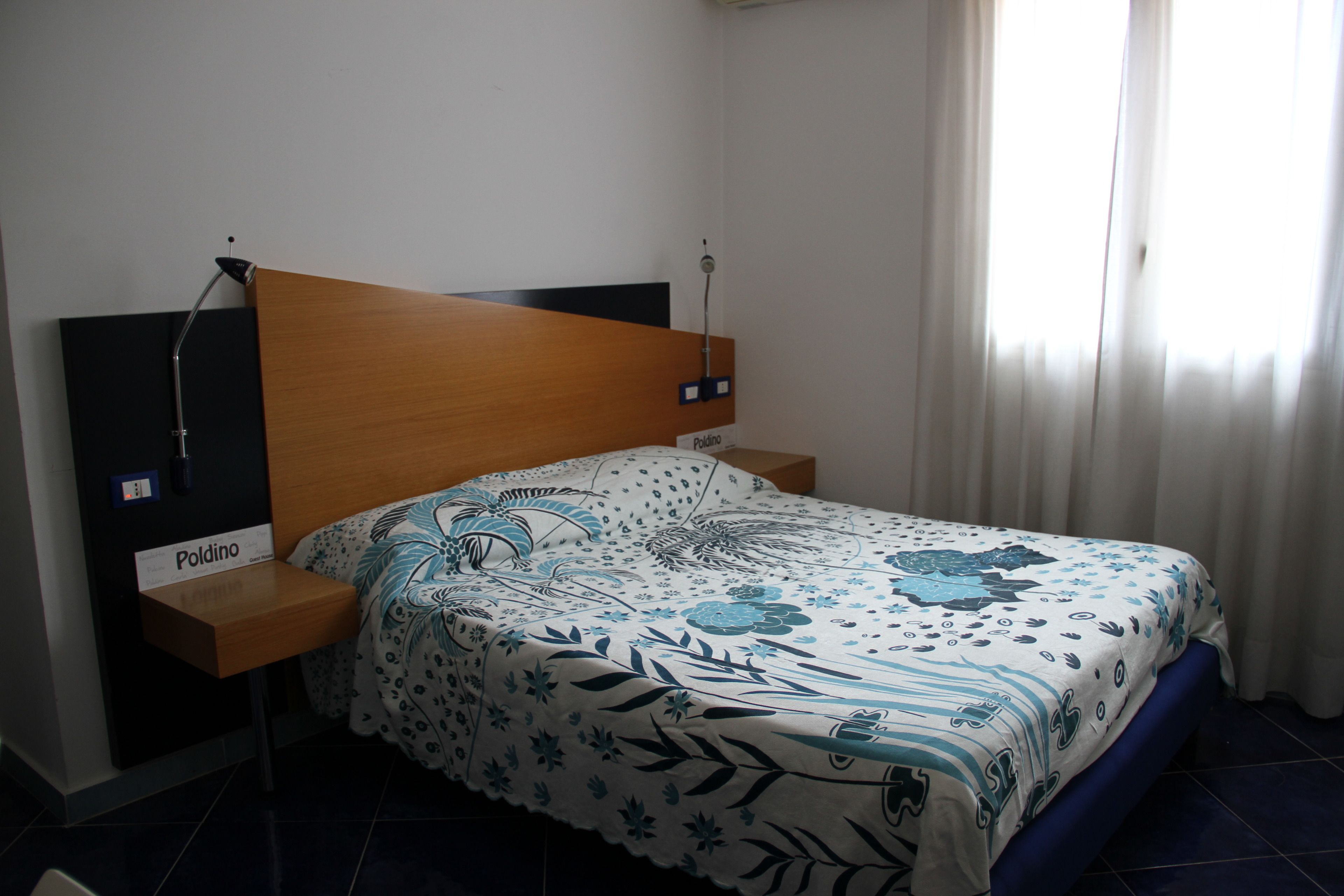 Quarto com cama, mesa de cabeceira, cabeceira de madeira, luminária e janela com cortinas.