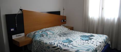 Quarto com cama, mesa de cabeceira, cabeceira de madeira, luminária e janela com cortinas.
