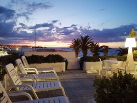 Terraço com cadeiras de descanso brancas e uma mesa, com vista para o pôr do sol, palmeiras e um horizonte urbano distante.