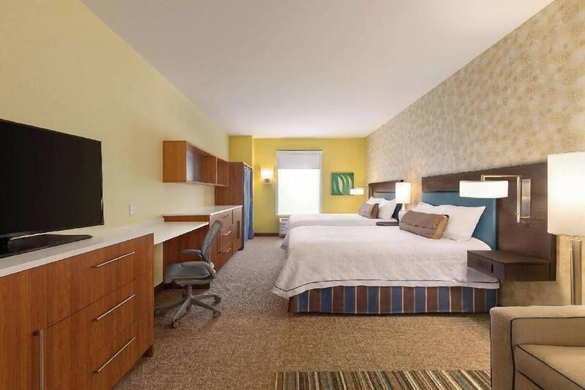 Quarto de hotel com cama, escrivaninha, cadeira, televisão e luminárias.