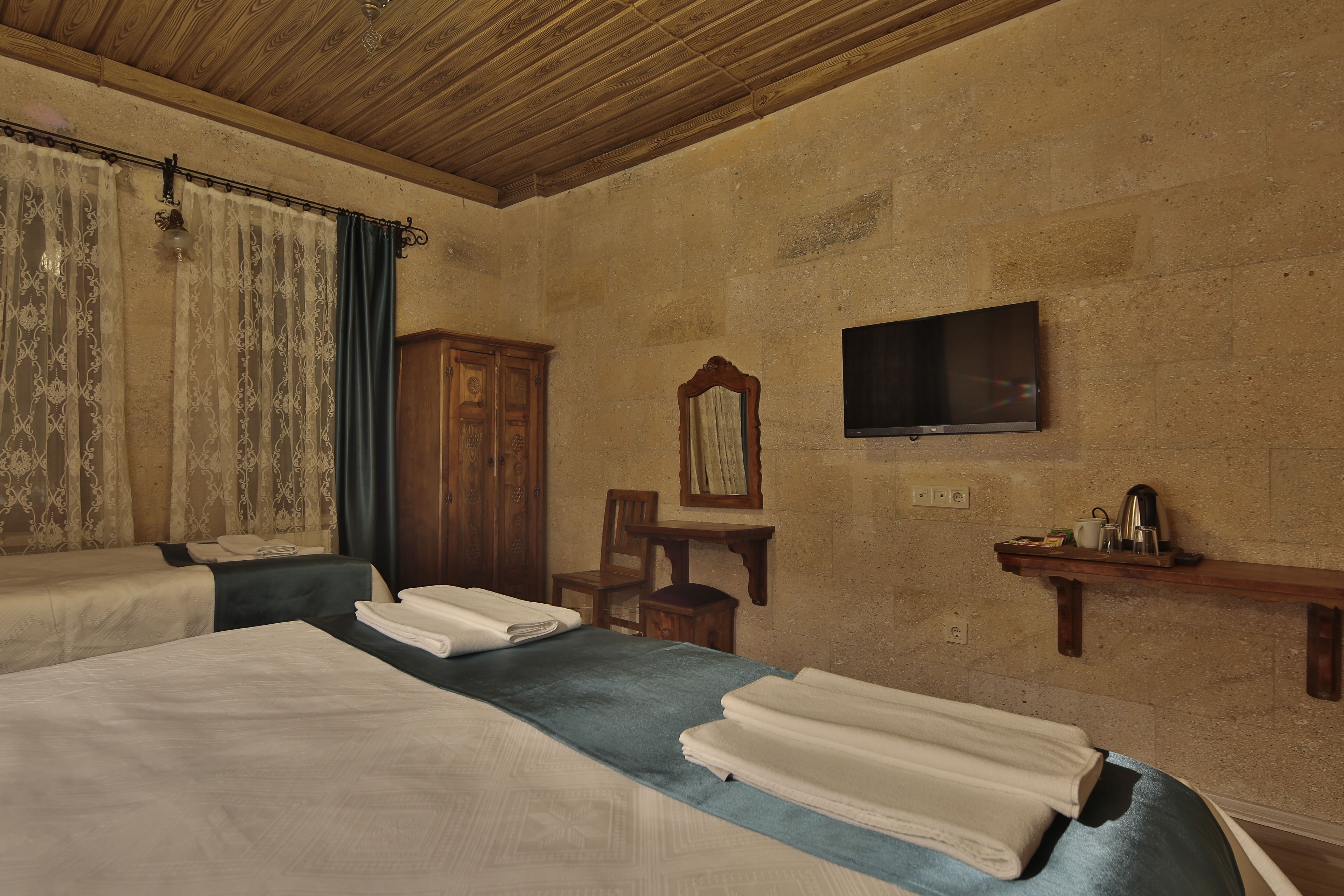 Quarto de hotel com duas camas, teto de madeira, televisão, armário de madeira e uma pequena mesa com uma cadeira.