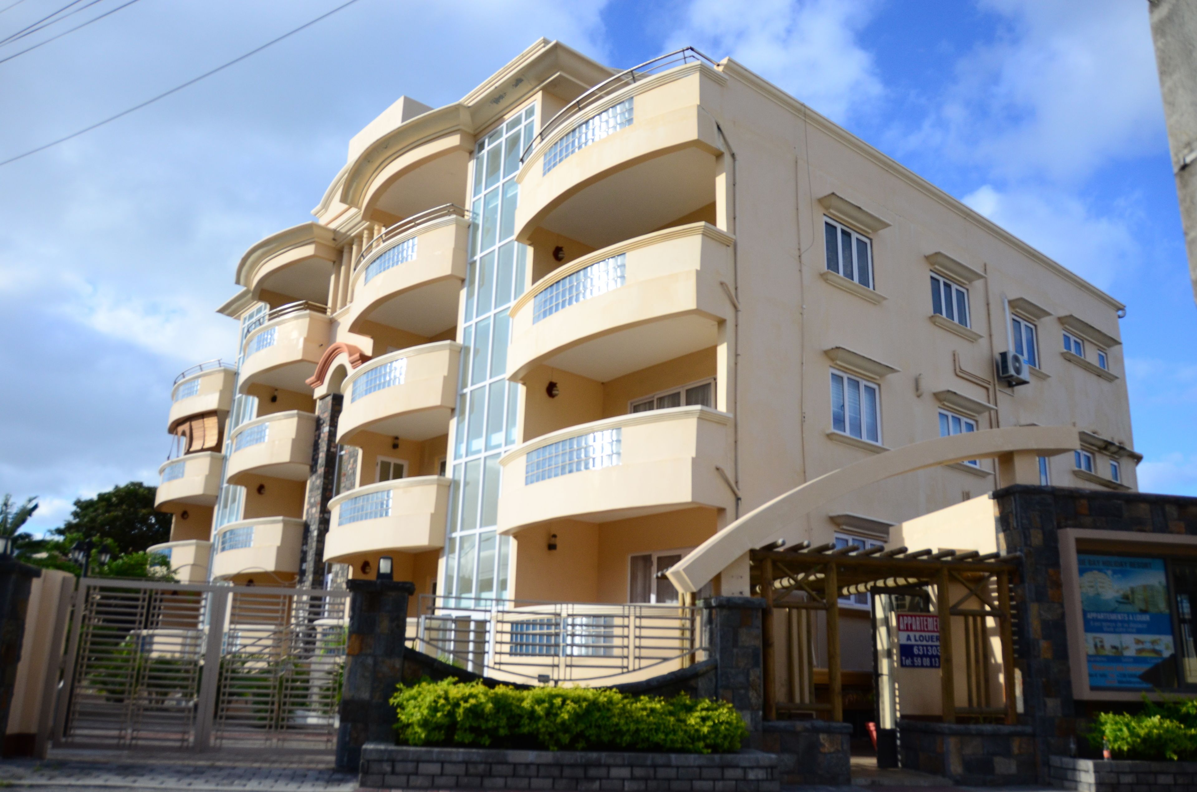 Um edifício de vários andares com varandas, entrada fechada por portão e placa indicando ser um prédio de apartamentos.
