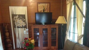 Uma sala de estar com piso de madeira, um sofá, uma TV em um armário, uma mesa de madeira com um vaso de flores e um abajur.