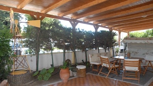 Uma área de varanda coberta com teto de madeira, mesa, cadeiras, plantas em vasos e um grande cachepot.