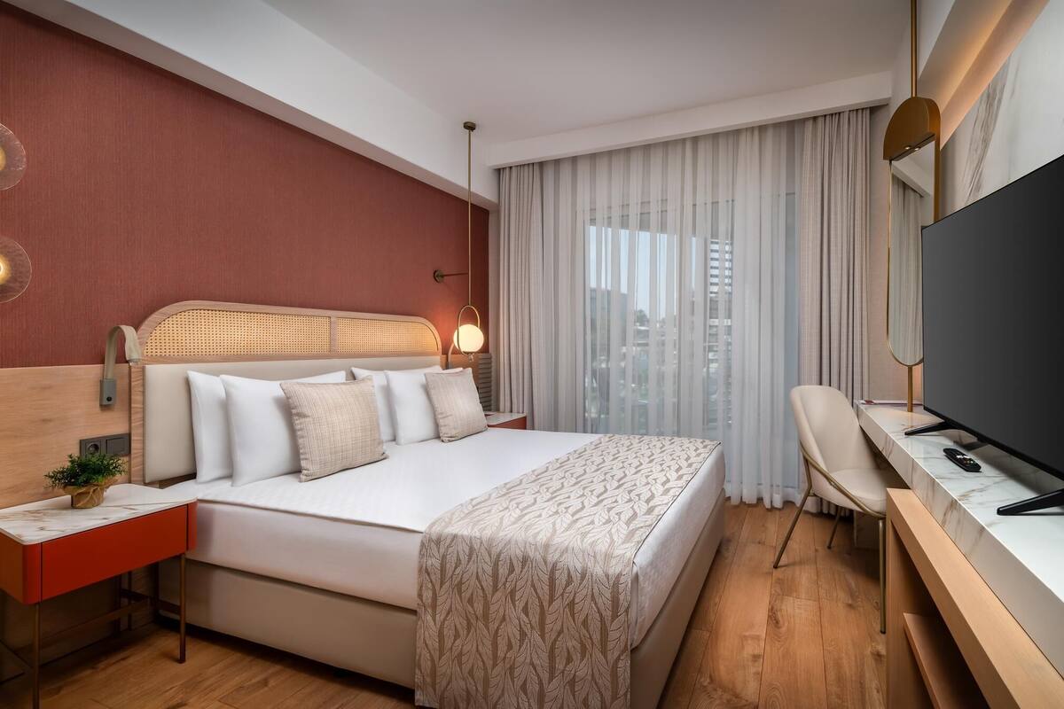 Quarto de hotel moderno com uma cama grande, uma mesa de escritório com televisão e vista para a cidade pela janela.