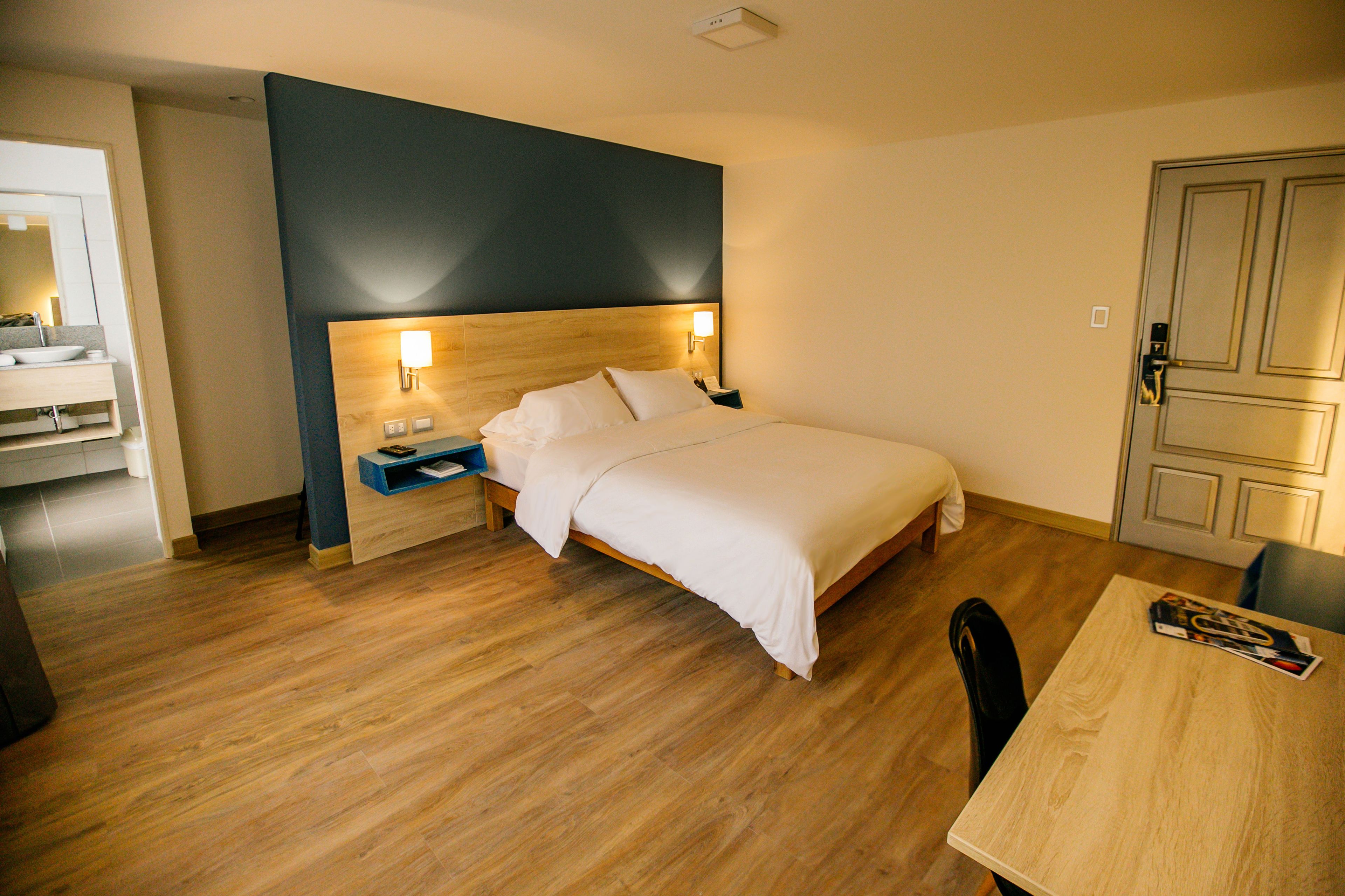 Quarto de hotel com piso de madeira, cama com roupa branca, mesa de cabeceira com abajur, escrivaninha com cadeira e porta.