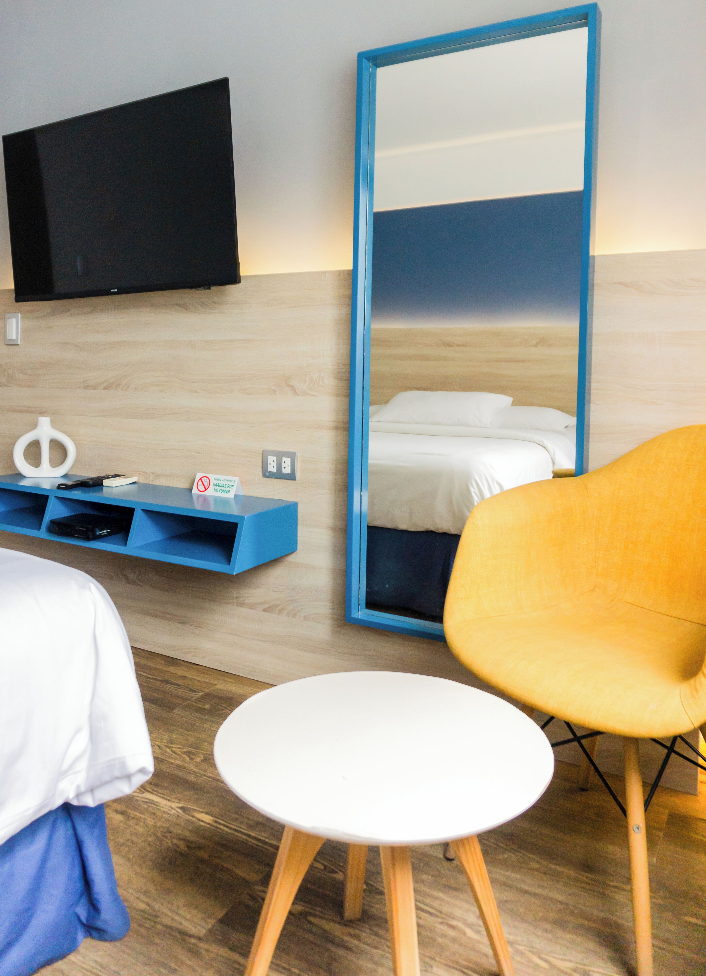 Quarto de hotel com cama, televisão na parede, espelho grande, mesa pequena e cadeira amarela.