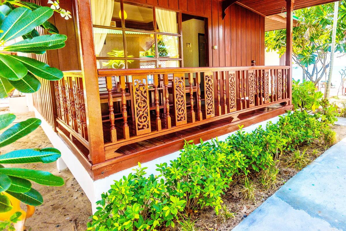 Varanda de madeira com corrimão, janela com cortinas e pequeno jardim com plantas verdes.