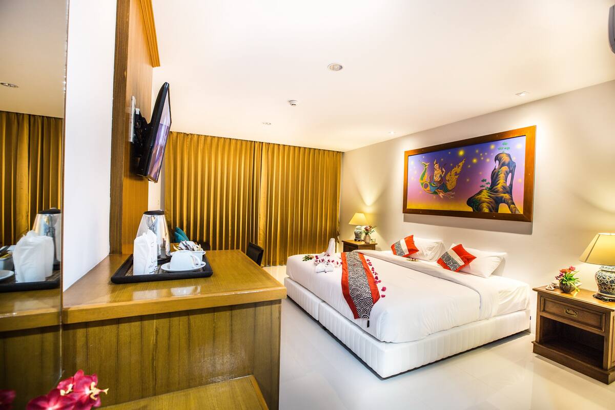 Quarto de hotel com cama, escrivaninha, televisão, quadro e mesa de cabeceira com abajur.