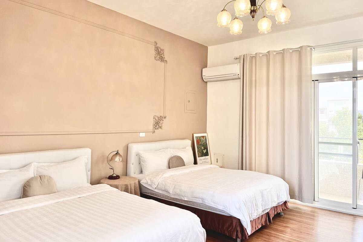 Quarto de hotel com duas camas, um candelabro, ar condicionado e uma janela grande com cortinas.