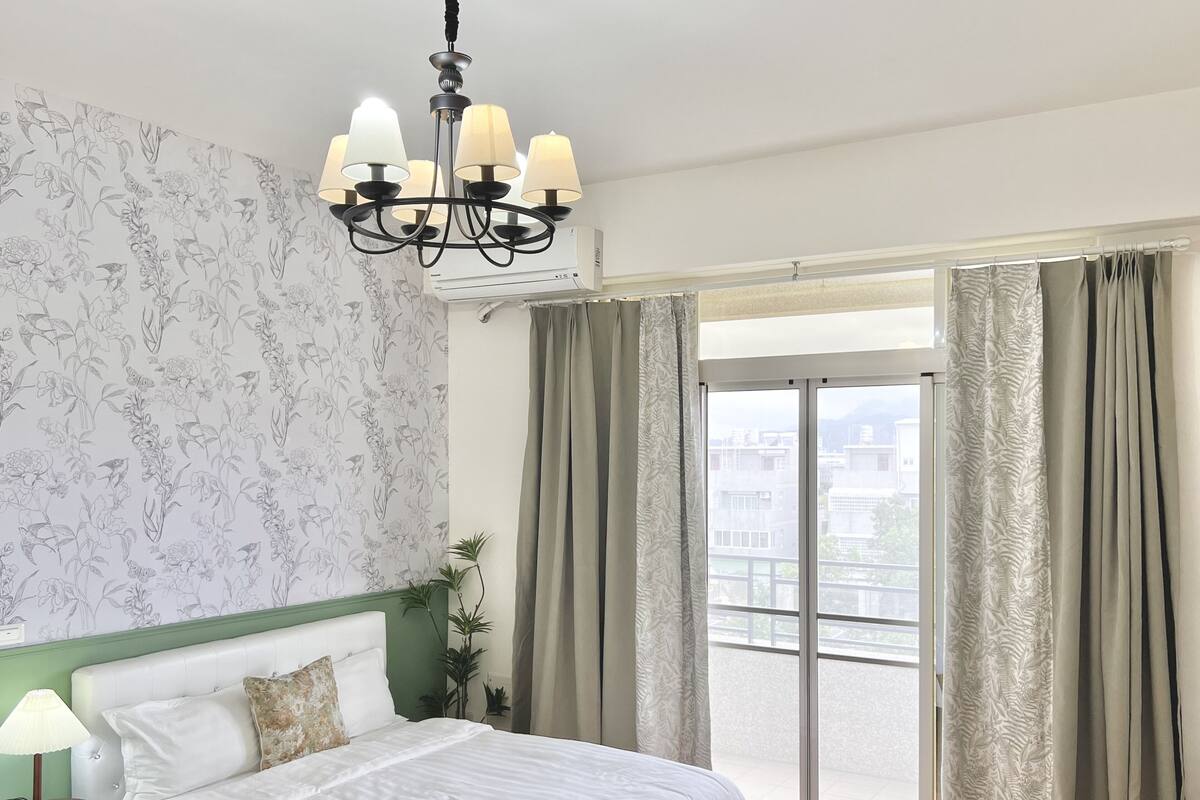 Quarto com cama, candelabro, janela com cortinas e vista para prédios.