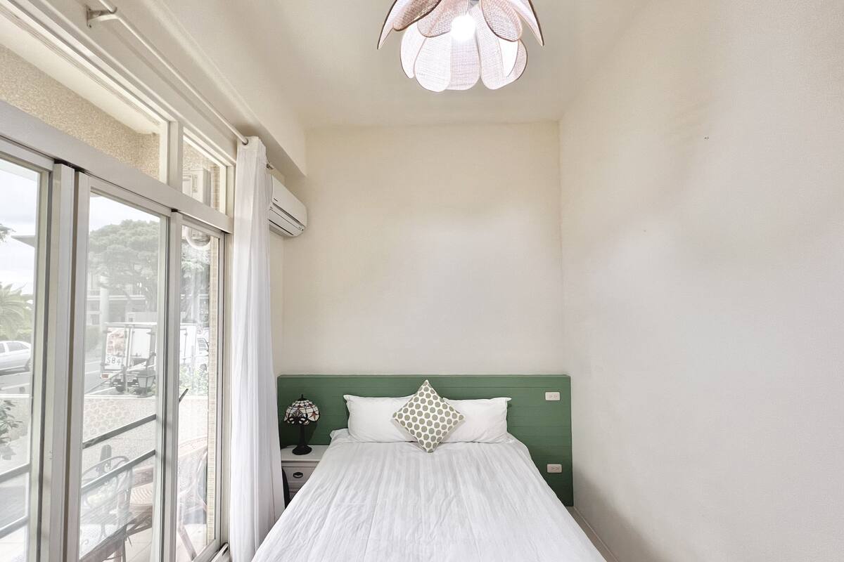 Uma cama pequena e bem arrumada com cabeceira verde em um quarto com uma janela grande.