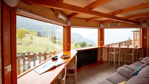Uma cabana de madeira com área de refeições, sofá e janelas amplas com vista para uma paisagem montanhosa.