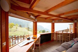 Uma cabana de madeira com área de refeições, sofá e janelas amplas com vista para uma paisagem montanhosa.