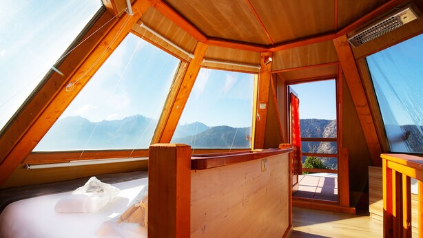 Quarto amplo com pé-direito alto, vigas de madeira e grandes janelas com vista para as montanhas.