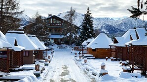Uma trilha coberta de neve que leva a um chalé de madeira com várias cabanas, cercado por montanhas nevadas.