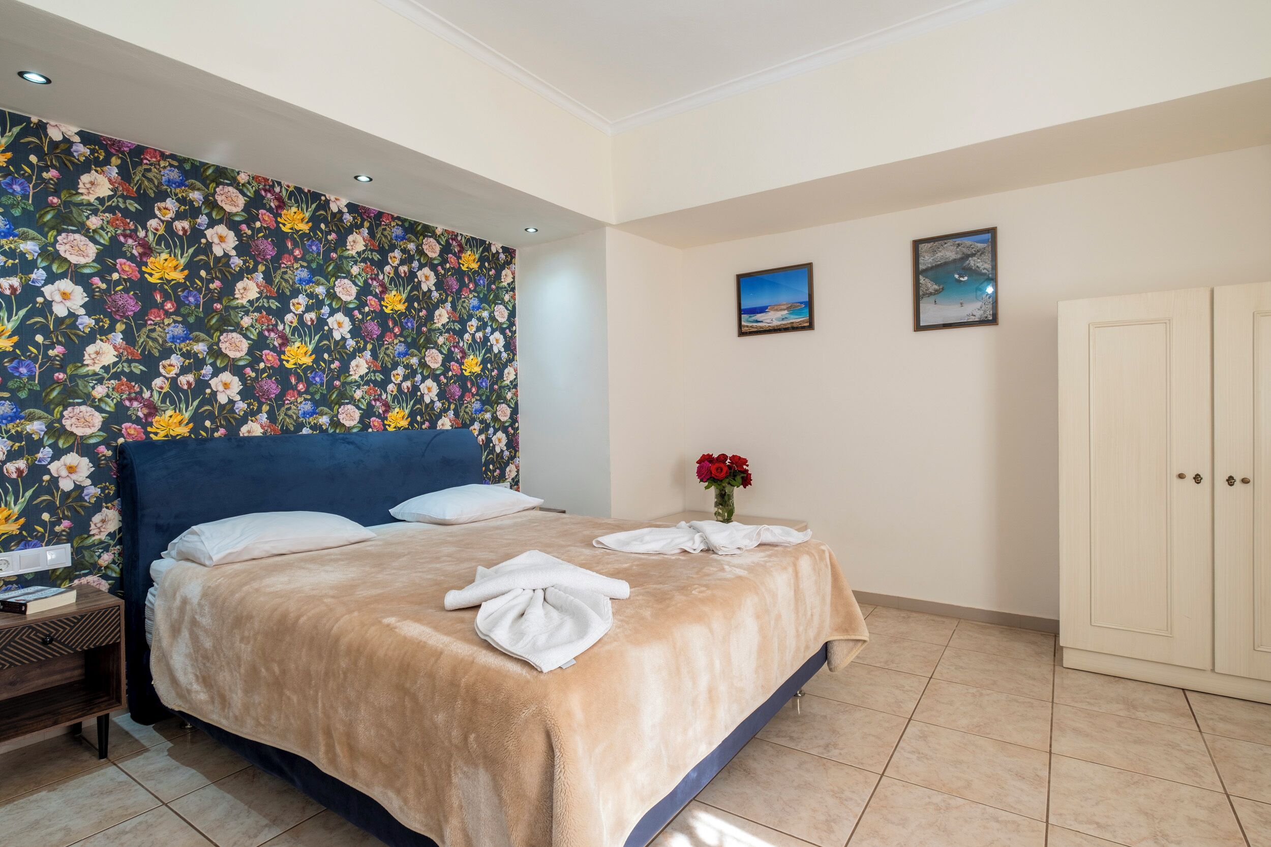 Uma cama bem arrumada com cabeceira floral, mesa de cabeceira com abajur e vaso com flores vermelhas na parede.