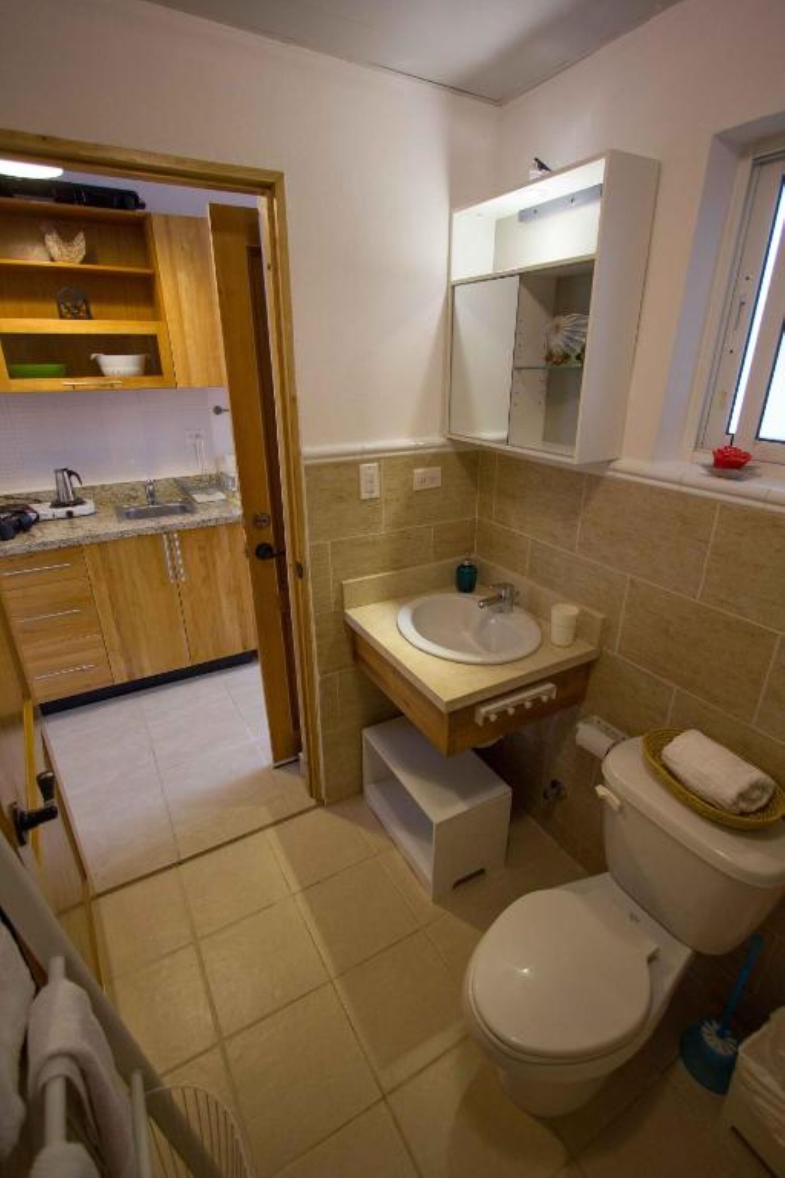 Casa de banho compacta com sanita, lavatório e espelho. Pequena janela acima do lavatório e porta que dá para uma área de cozinha.