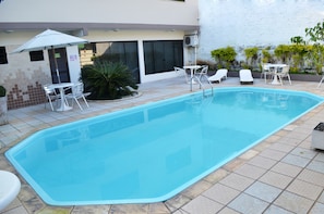 Piscina com deck ladrilhado, rodeada por cadeiras de lounge brancas e uma pequena mesa.
