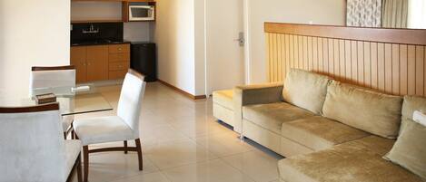 Uma sala moderna com sofá, mesa de jantar com cadeiras, área de cozinha com armários e micro-ondas, e uma porta que dá para outro cômodo.