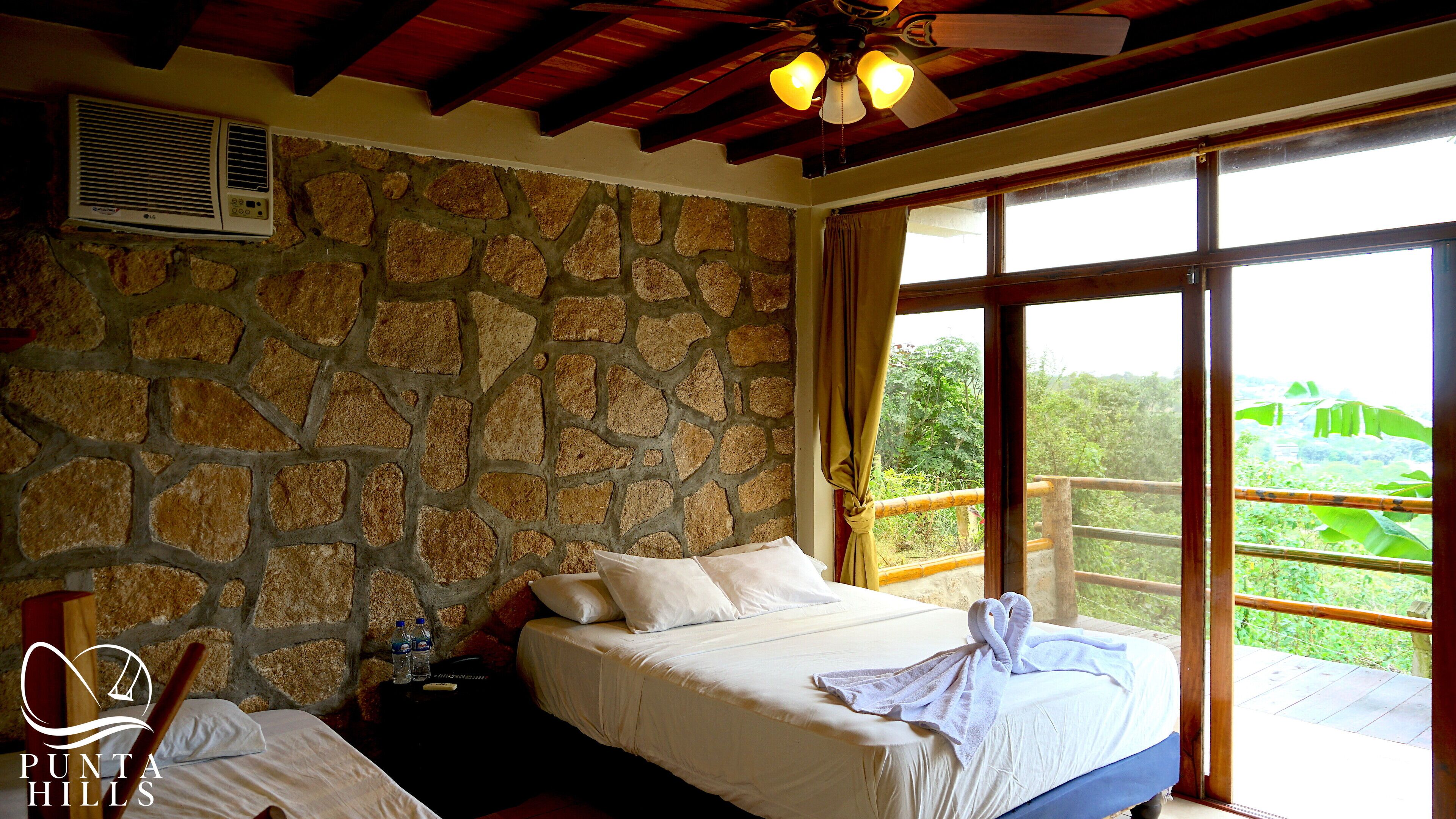 Quarto com parede de pedra, cama com roupa de cama branca, ventoinha no teto e janela com vista para vegetação.