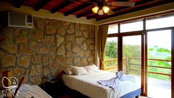 Quarto com parede de pedra, cama com roupa de cama branca, ventoinha no teto e janela com vista para vegetação.