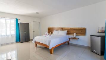 Quarto de hotel moderno com uma cama grande, cabeceira de madeira, mesa de cabeceira pequena, frigorífico e casa de banho com chuveiro e sanita.