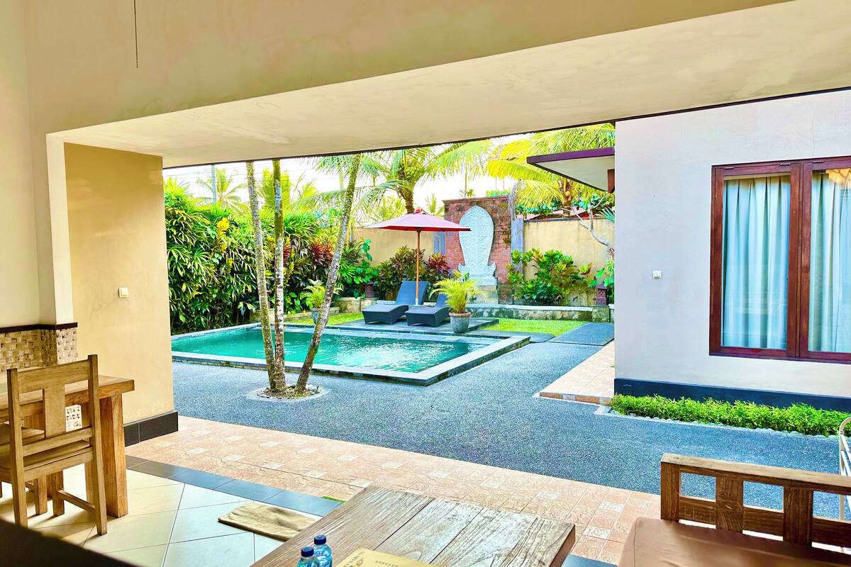 Uma área de piscina com mesa e cadeiras de madeira, cercada por vegetação exuberante e uma pequena estátua.