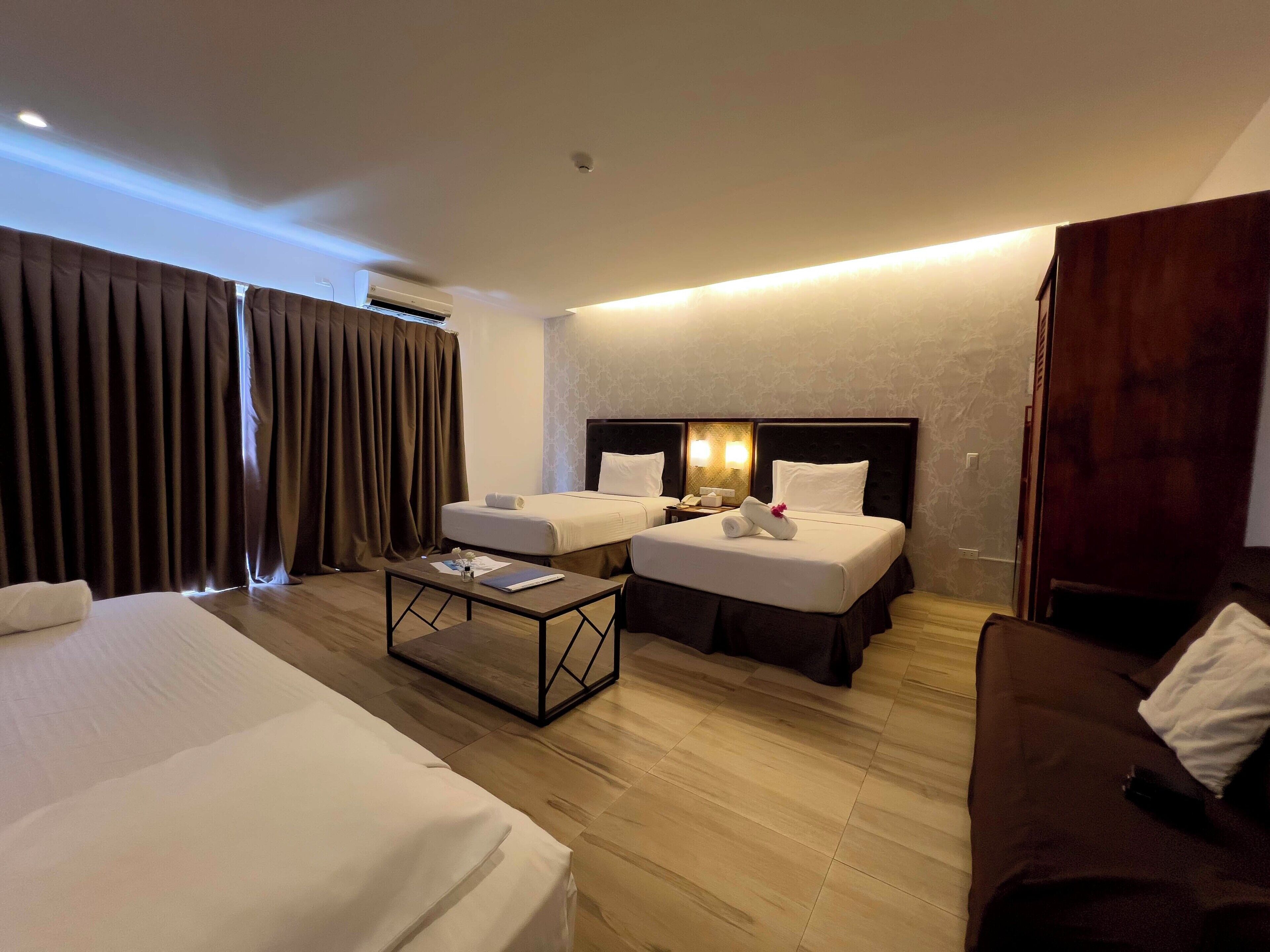 Quarto de hotel com duas camas, um sofá, uma mesa de centro e uma janela grande com cortinas.