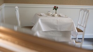 Mesa de jantar posta com toalha branca, pratos e um vaso com flores vermelhas.