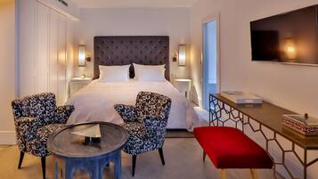 Quarto de hotel com uma cama grande, duas poltronas, uma mesa redonda pequena, um banco vermelho, uma televisão e uma mesa de cabeceira com abajur.