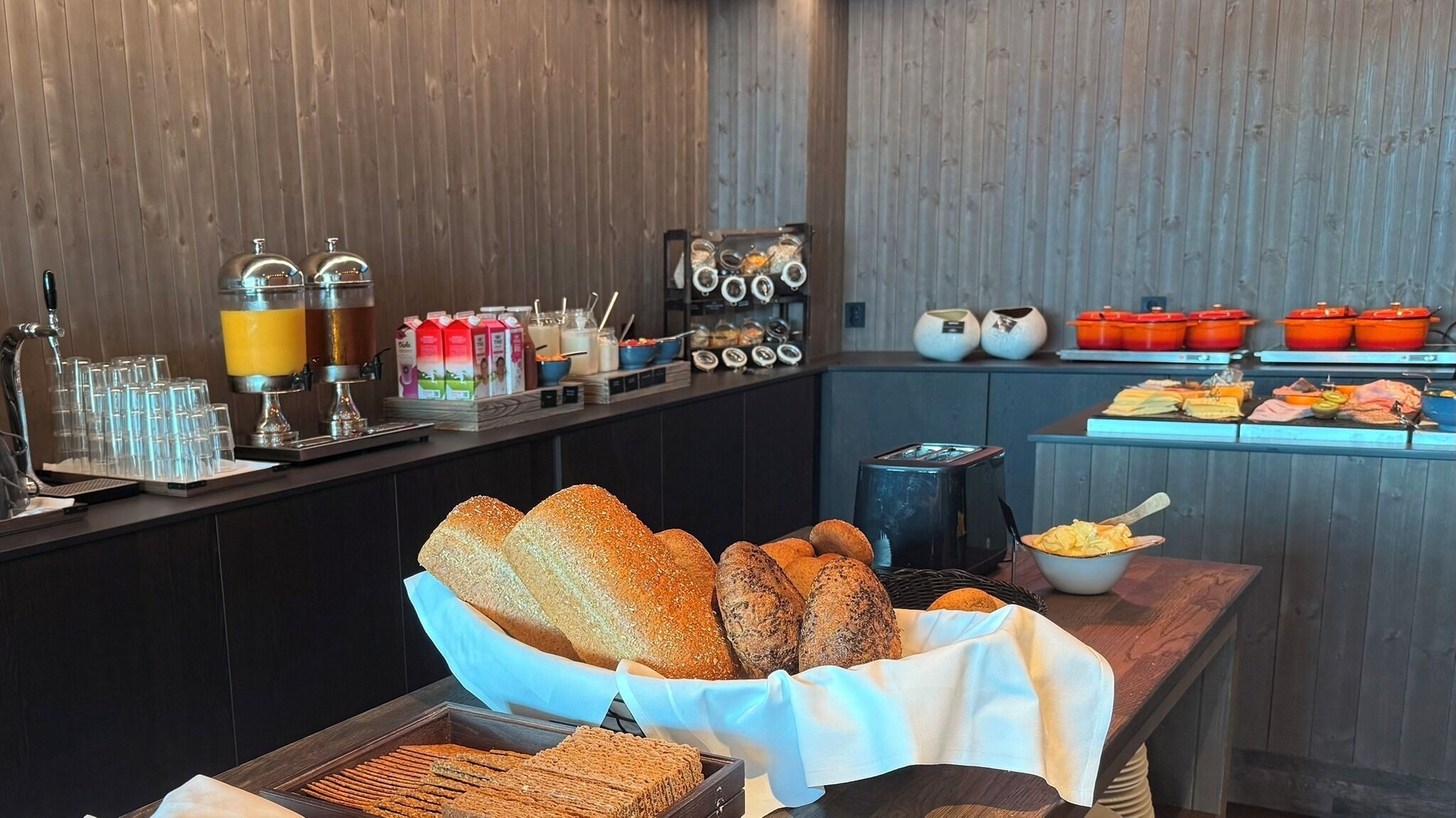 Uma mesa de buffet com diversos pães, um dispensador de suco e uma torradeira.
