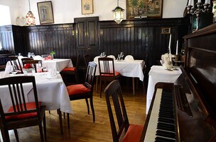 Uma sala de jantar com mesas preparadas para dois, cadeiras de madeira, um piano e lustres fixados na parede.
