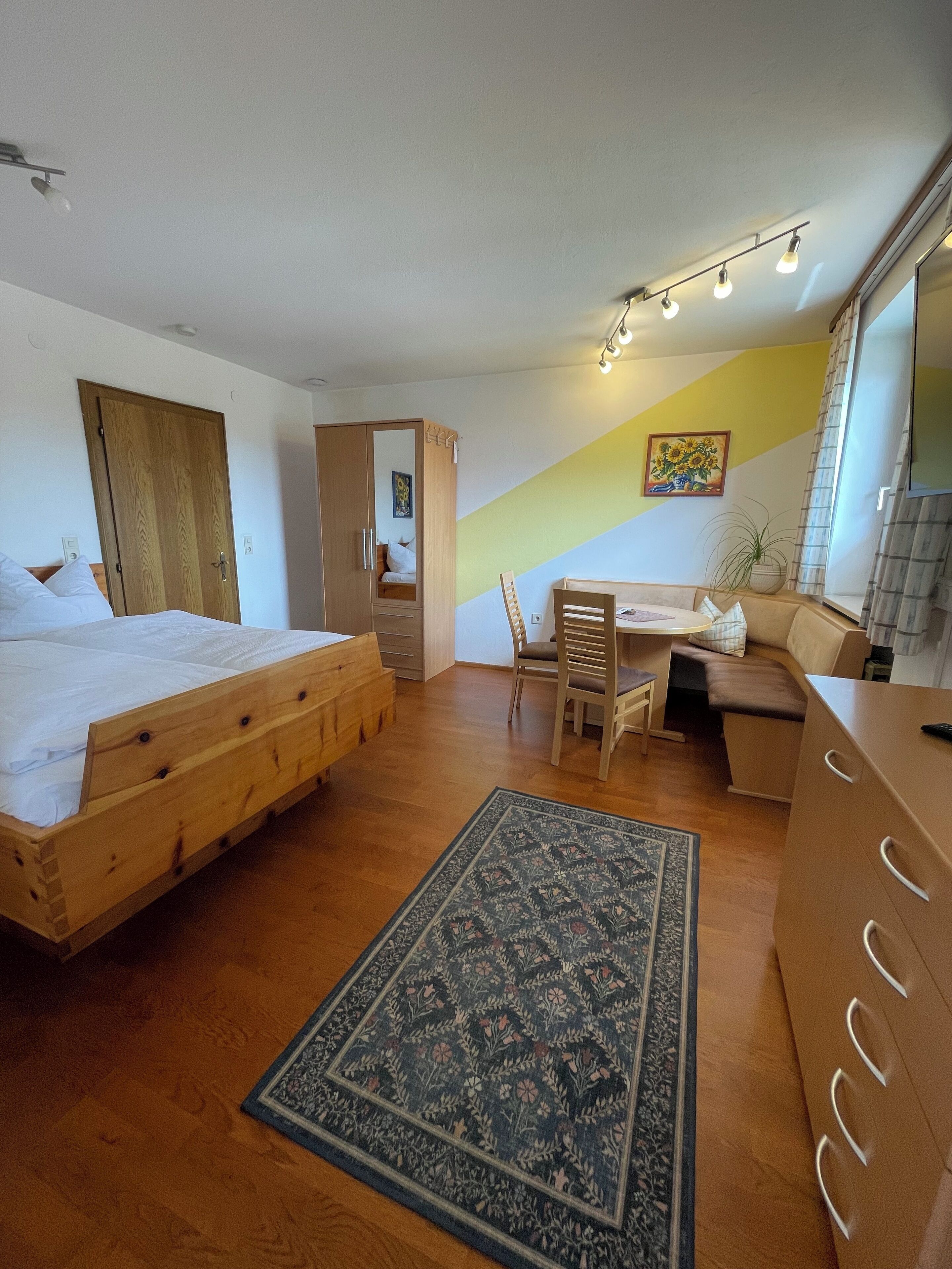 Quarto com cama de madeira, uma escrivaninha e um sofá. Há um espelho, uma planta e um tapete.
