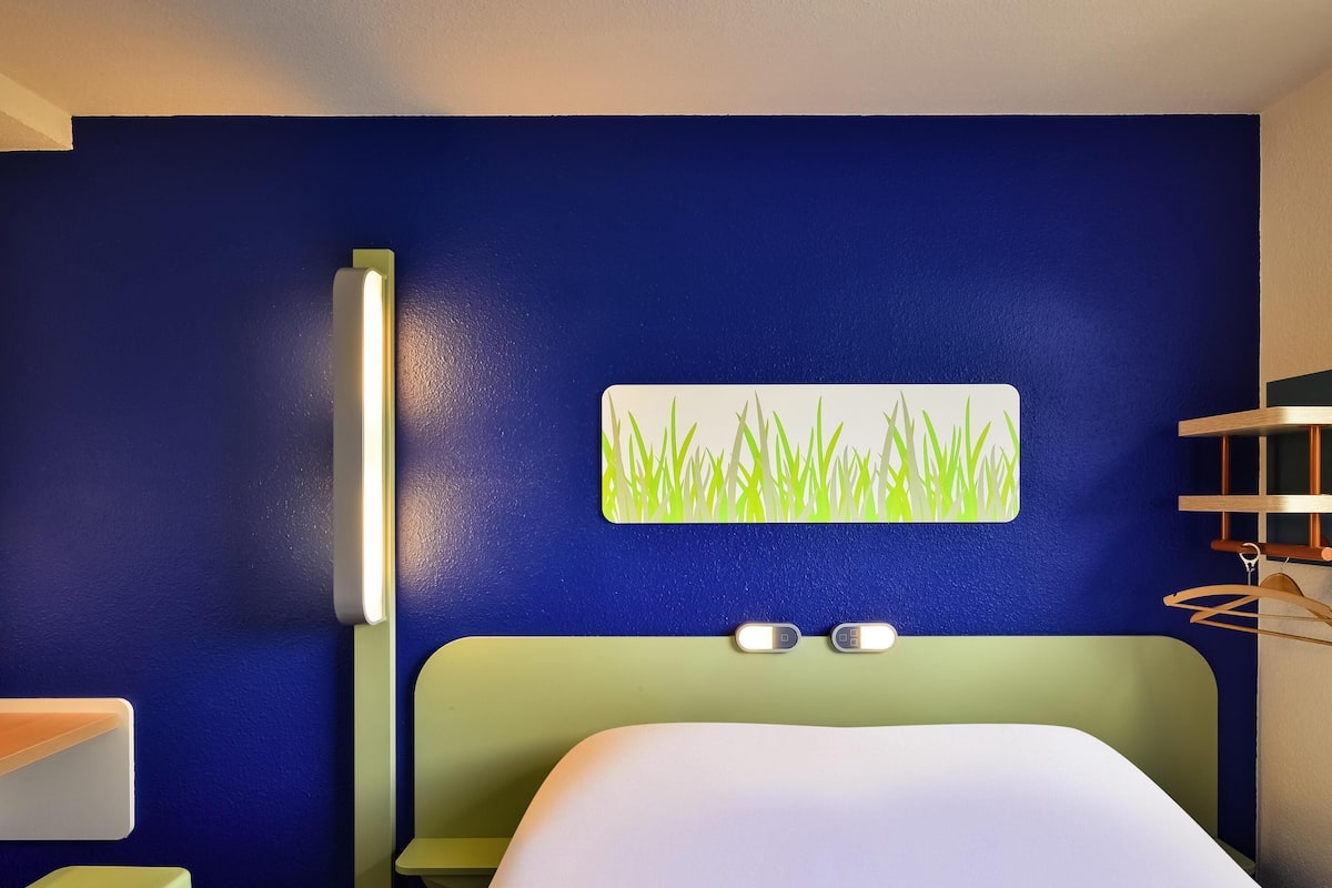 Quarto de hotel com cama, luminária na parede, uma pequena mesa e uma obra de arte na parede.