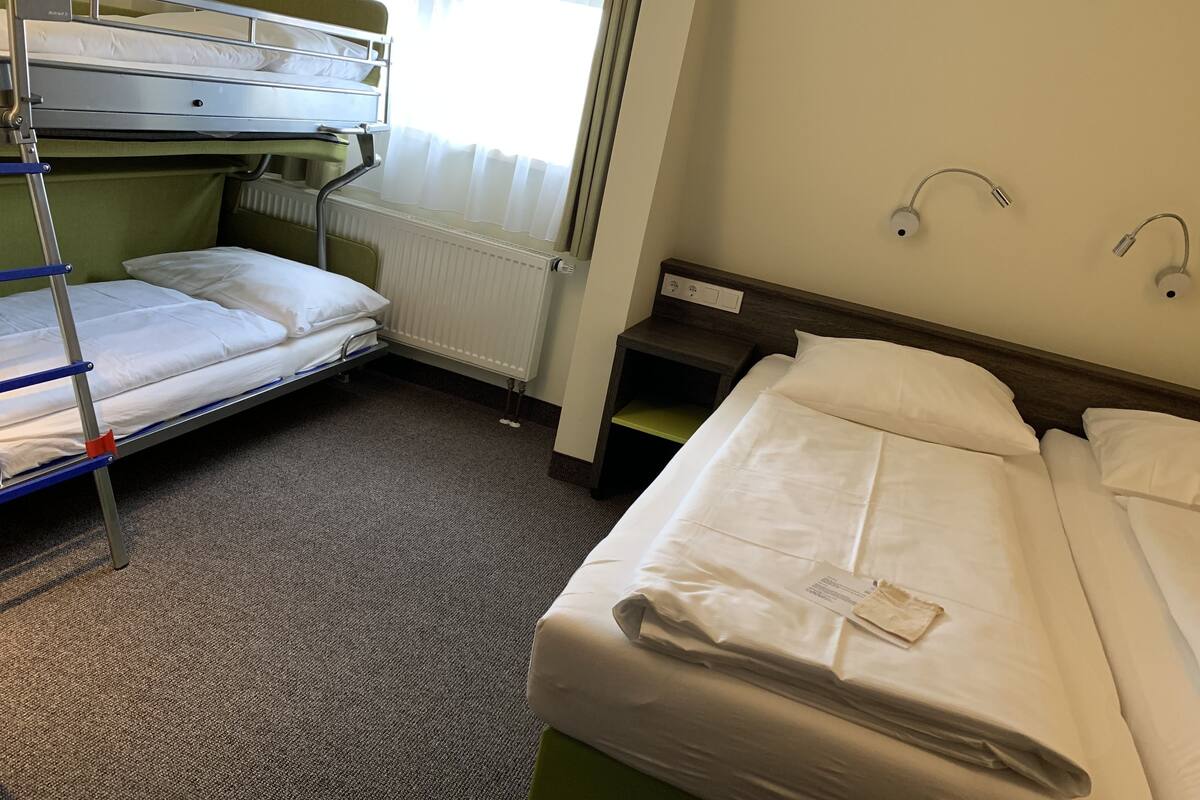 Quarto de hotel com beliche, uma cama de solteiro, mesa de cabeceira, cortinas na janela e radiador.