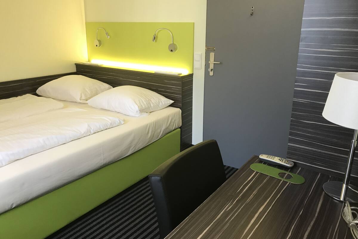 Quarto de hotel com uma cama, uma mesa, uma cadeira, um abajur e uma porta.