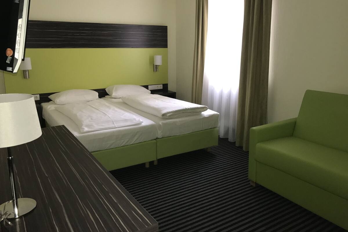 Quarto de hotel com cama, secretária, poltrona verde e janela com cortinas.