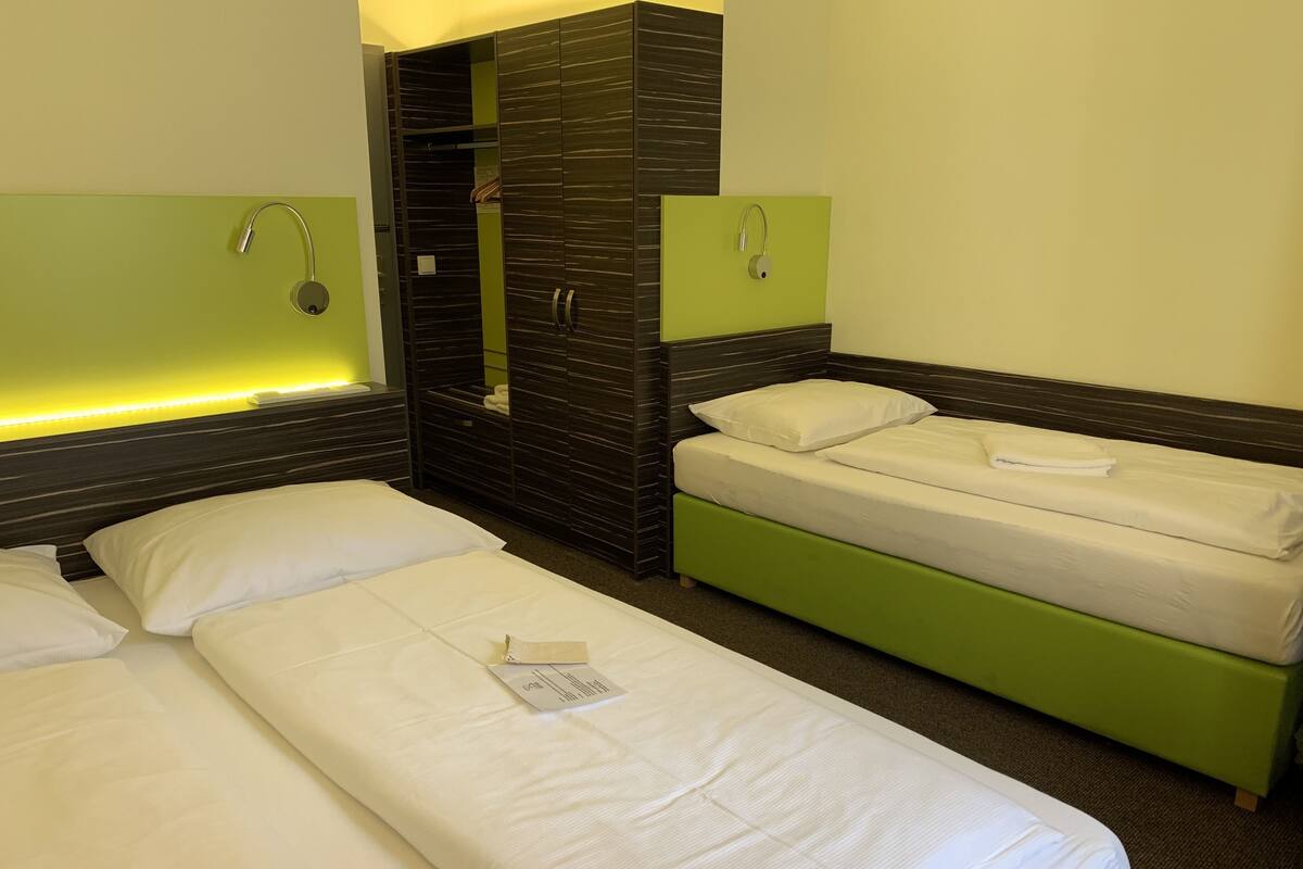 Quarto de hotel com duas camas, cada uma com roupa de cama branca e uma etiqueta no colchão. Possui cabeceira verde e um guarda-roupa de madeira escura.
