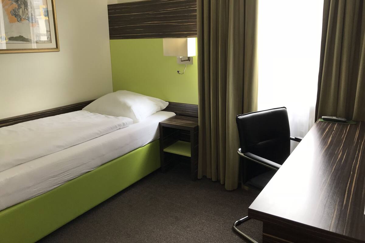 Quarto de hotel com uma cama de solteiro, uma escrivaninha, uma cadeira, um abajur e uma janela com cortinas.