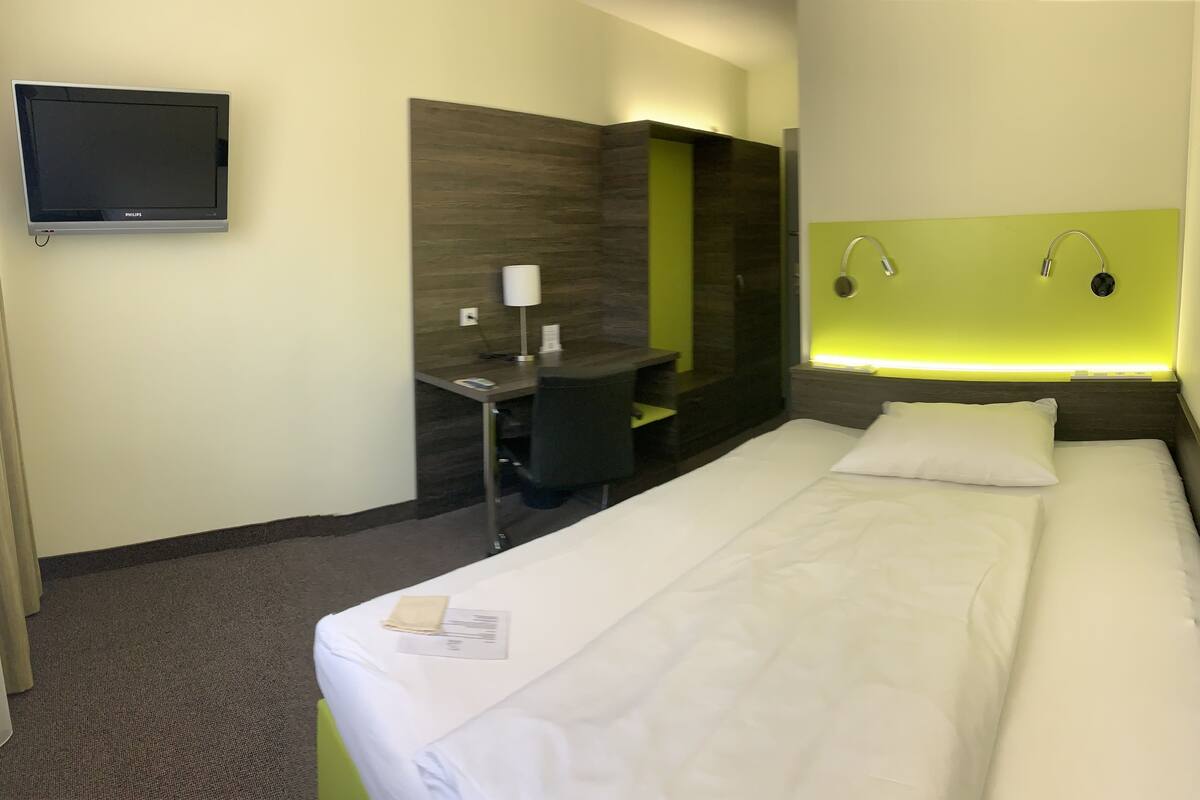 Quarto de hotel com cama, escrivaninha, cadeira, televisão de tela plana e luminária na parede.