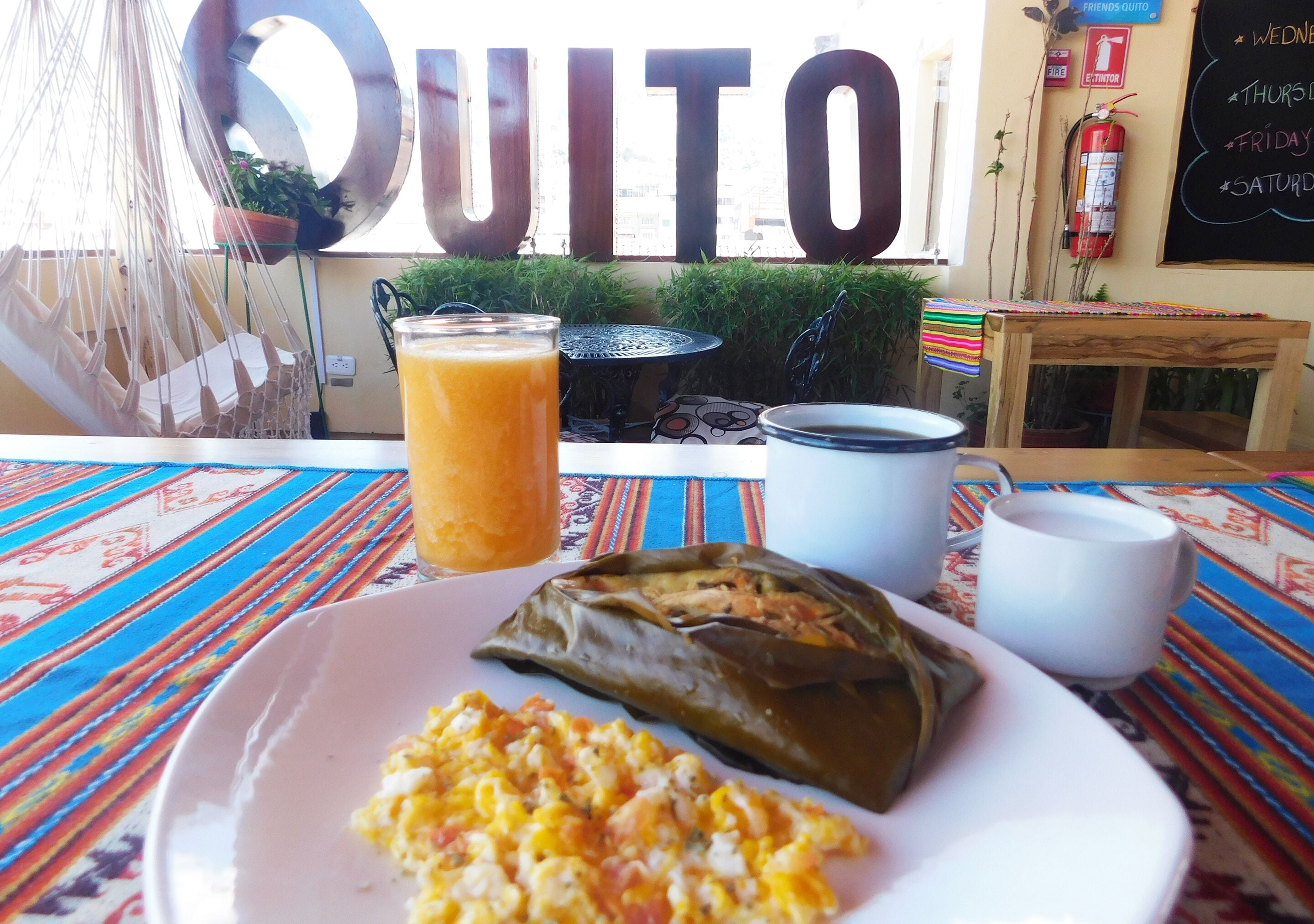 Uma refeição com um prato de comida, um copo de suco de laranja e uma xícara branca em cima de uma mesa com toalha colorida.