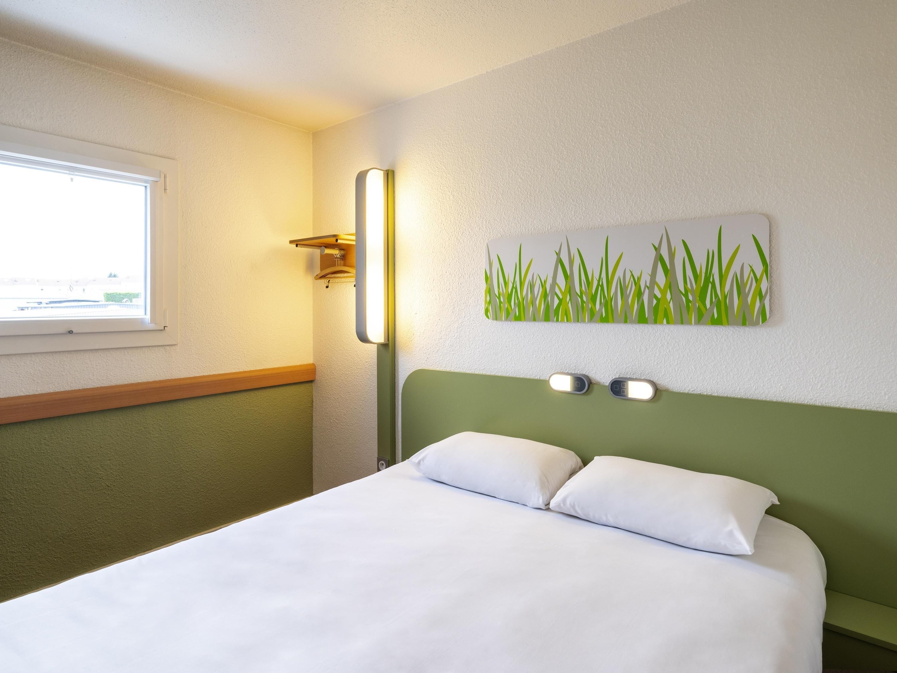 Quarto de hotel com cama, janela, luminária na parede e pintura de relva.