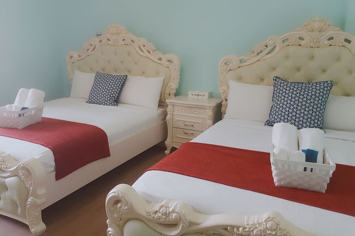 Duas camas com cabeceiras elaboradas, um cobertor vermelho, uma mesa de cabeceira branca e um cesto com artigos de higiene pessoal.