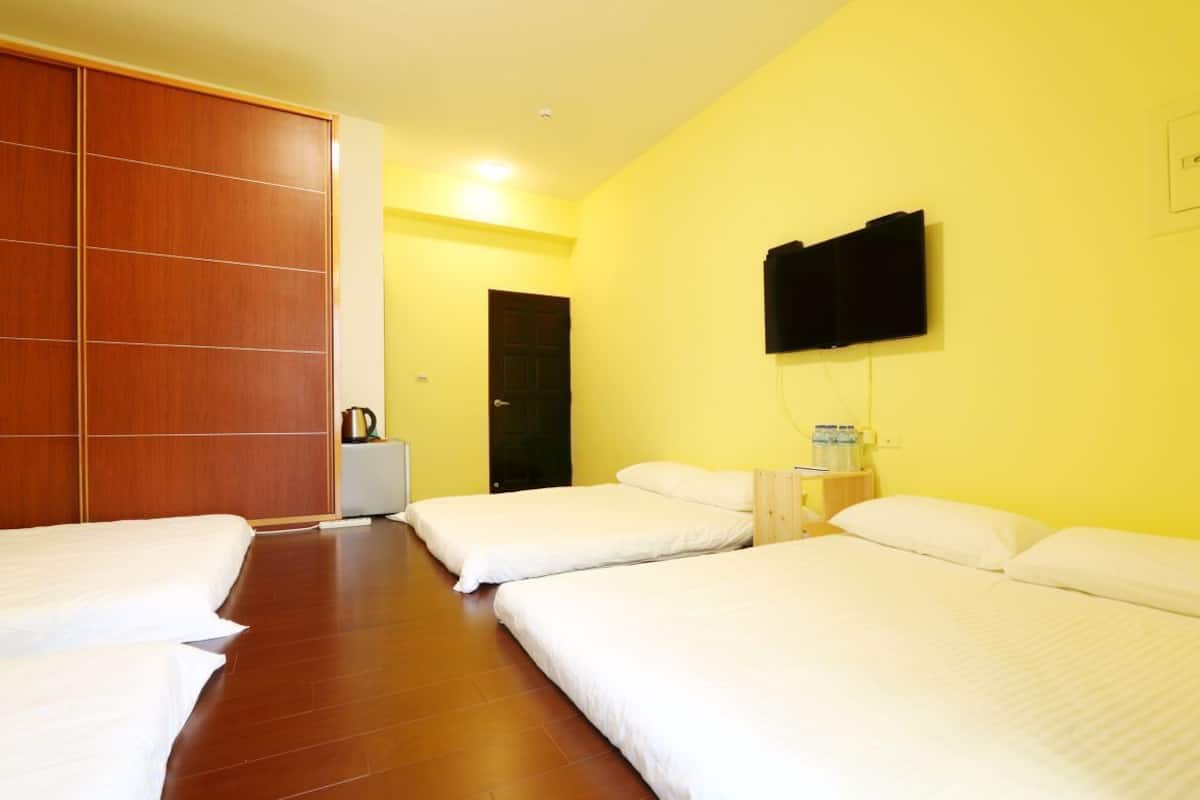 Quarto de hotel com três camas de solteiro, um guarda-roupa de madeira, televisão de tela plana na parede e uma mesa de cabeceira com uma garrafa de água.