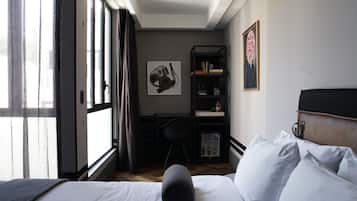 Quarto de hotel com cama, escrivaninha com cadeira, estante e janela com cortinas.