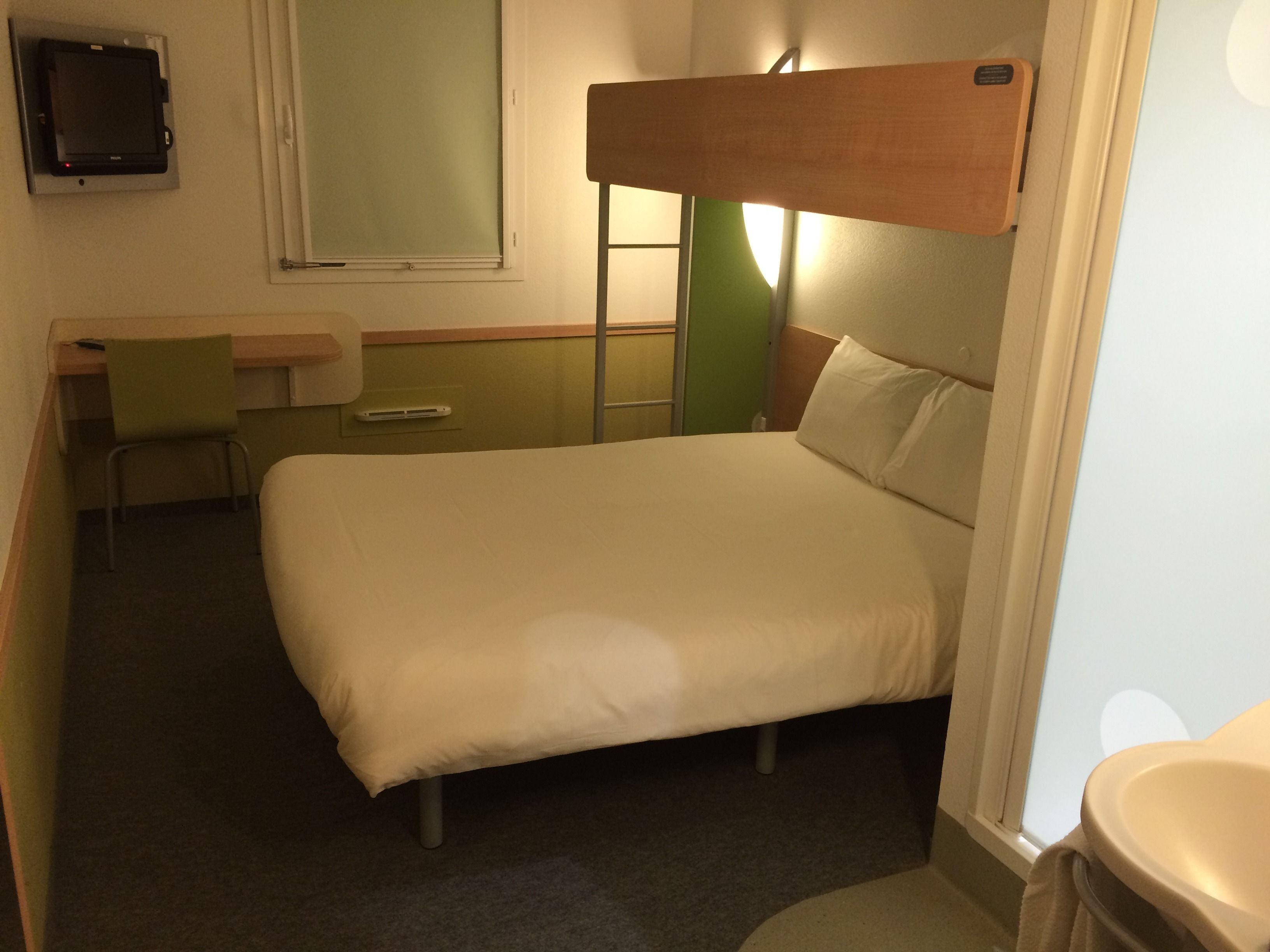 Quarto de hotel compacto com uma cama de solteiro, uma escrivaninha e uma cadeira.